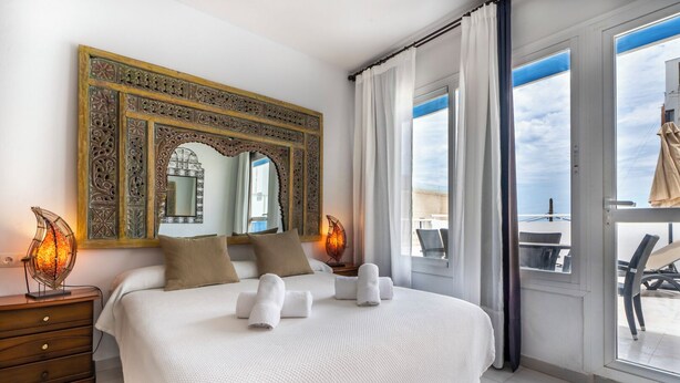 Quarto de hotel com uma cama grande, um espelho decorativo, duas luminárias de cabeceira e vista para a varanda externa.