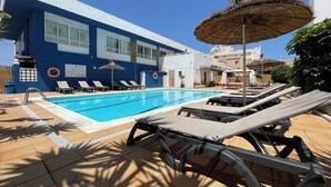 Uma área de piscina com espreguiçadeiras e guarda-sóis, céu limpo e um edifício com paredes azuis e brancas.