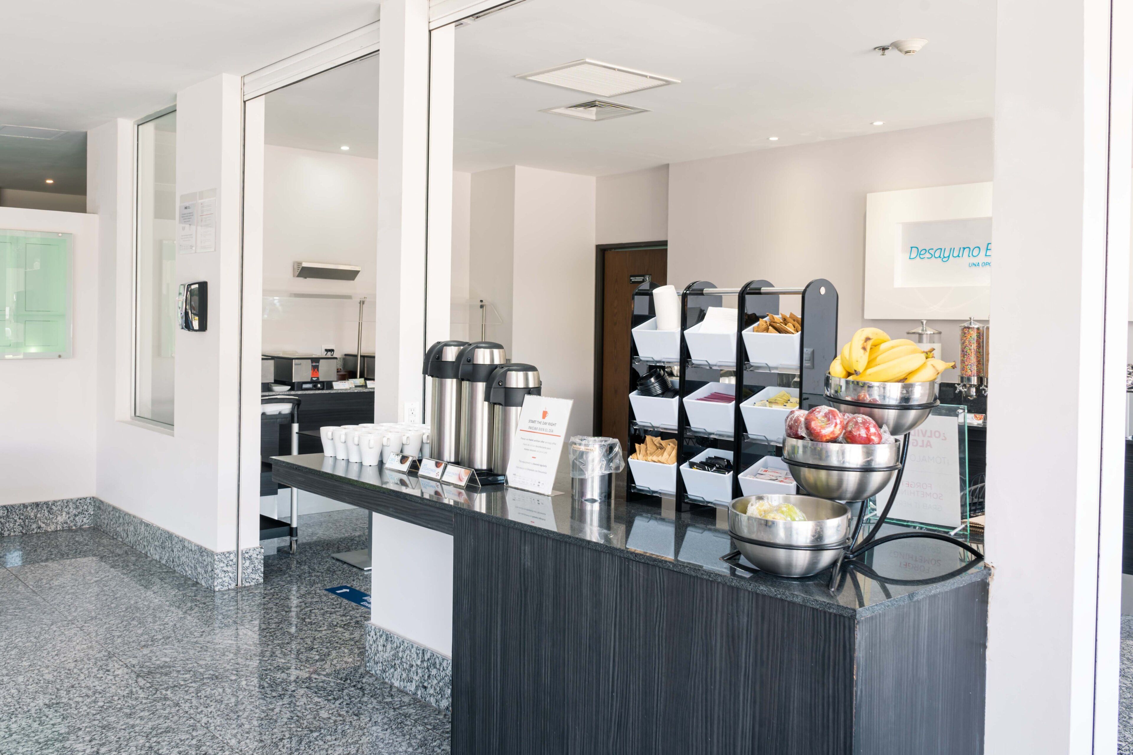 Uma área de recepção de hotel com um espaço para café, uma vitrine de vidro com frutas e uma placa para Desagüno.