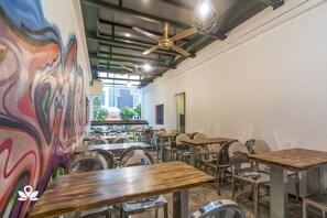 Uma área de refeições com várias mesas e cadeiras, ventiladores de teto e vista da cidade por grandes janelas.