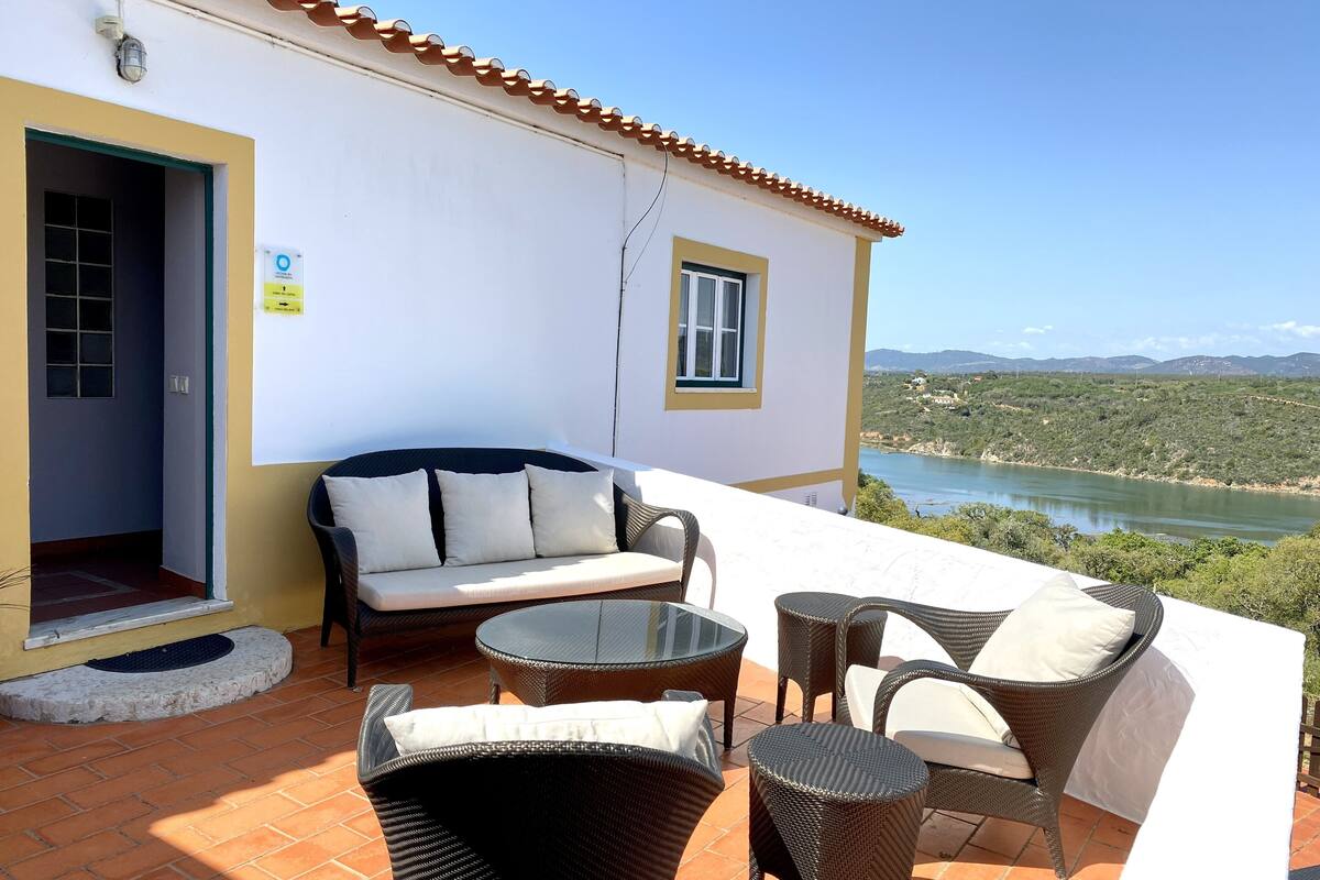 Terraço com móveis de vime, um sofá e uma mesa. Dá-se para um rio e colinas ao fundo.