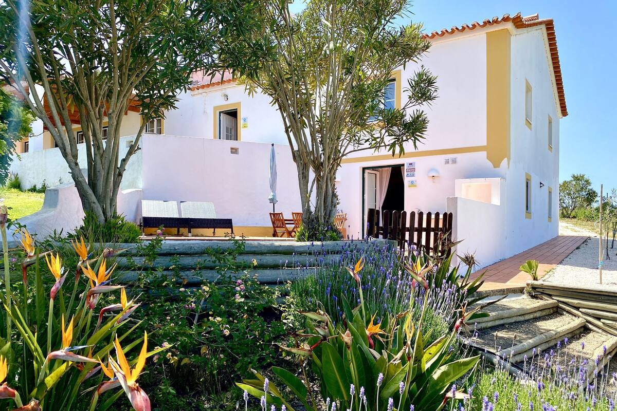 Um edifício branco com telhado vermelho, cerca de madeira e um jardim com diversas plantas e flores.