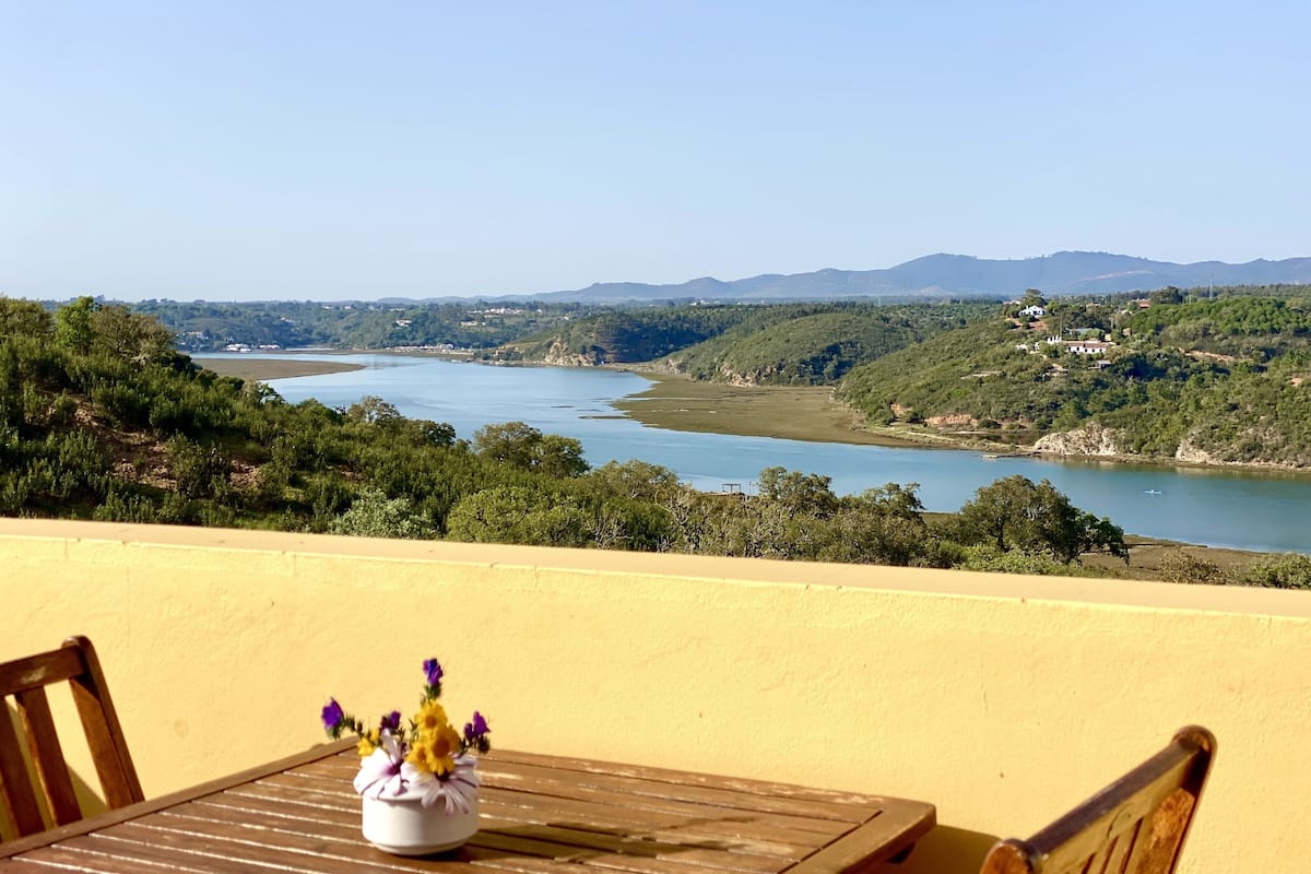 Uma varanda com mesa e cadeiras, com vista para um rio e colinas.