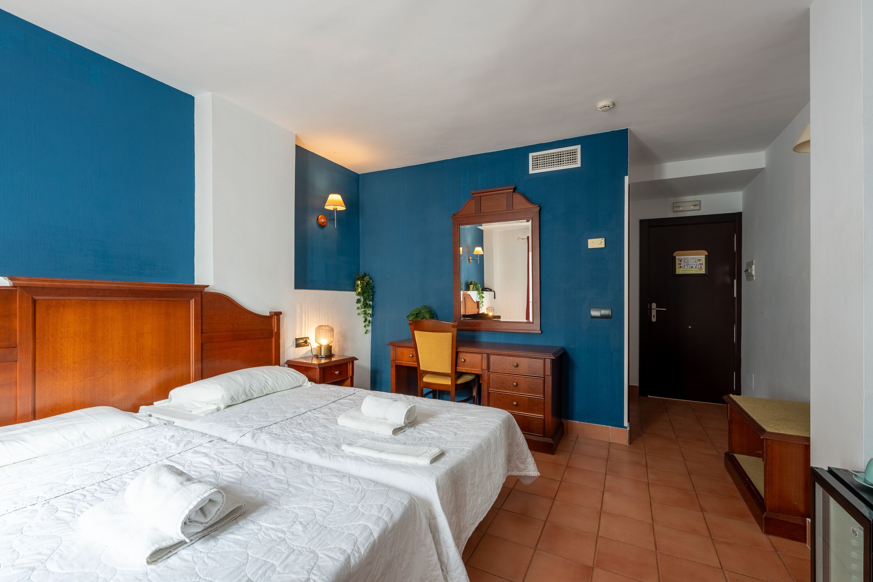 Quarto de hotel com uma cama grande, cómoda de madeira, escrivaninha e espelho.