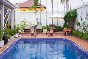 Piscina com azulejos azuis, duas espreguiçadeiras de madeira e uma mesa com guarda-sol branco.