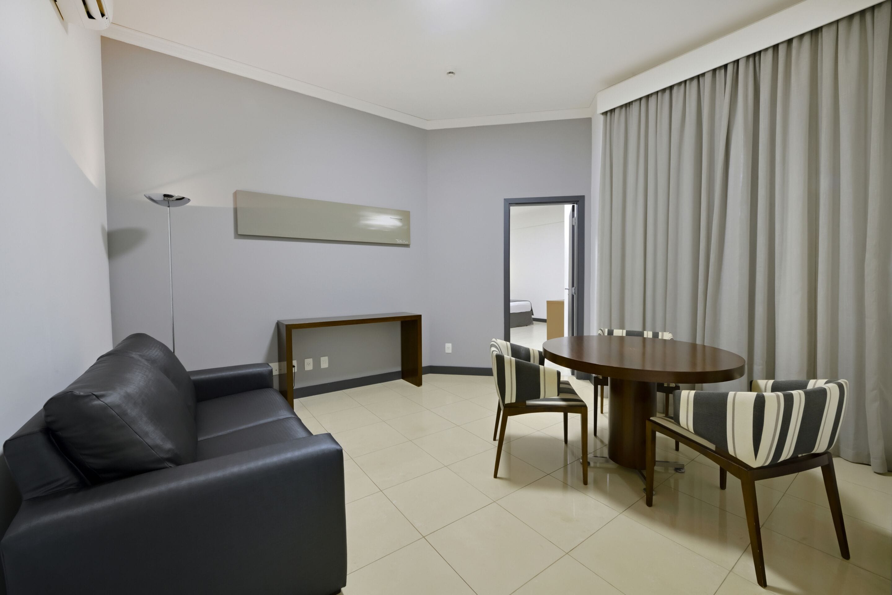 Uma sala de estar moderna com um sofá de couro escuro, uma mesa de jantar redonda com cadeiras, um abajur e um espelho grande na parede.