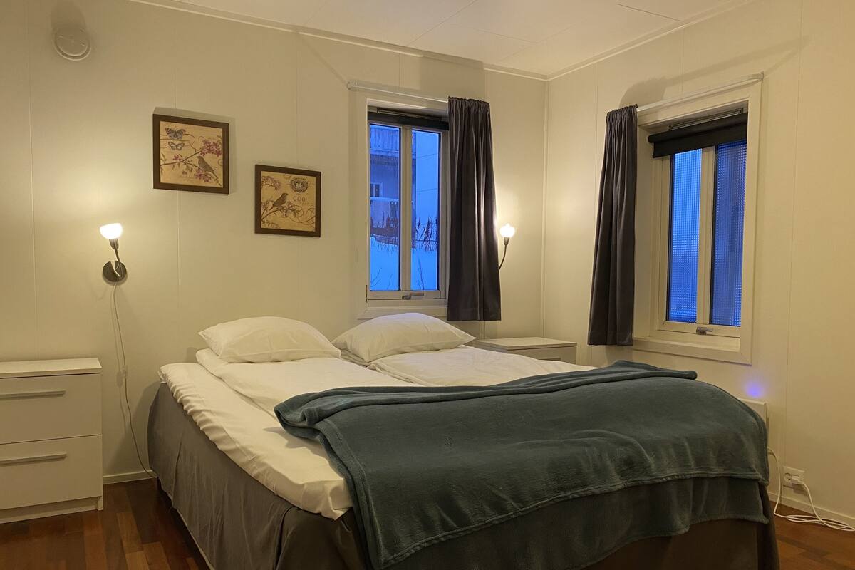 Quarto com uma cama, duas mesas de cabeceira, um criado-mudo, janela com cortinas e luminárias fixadas na parede.
