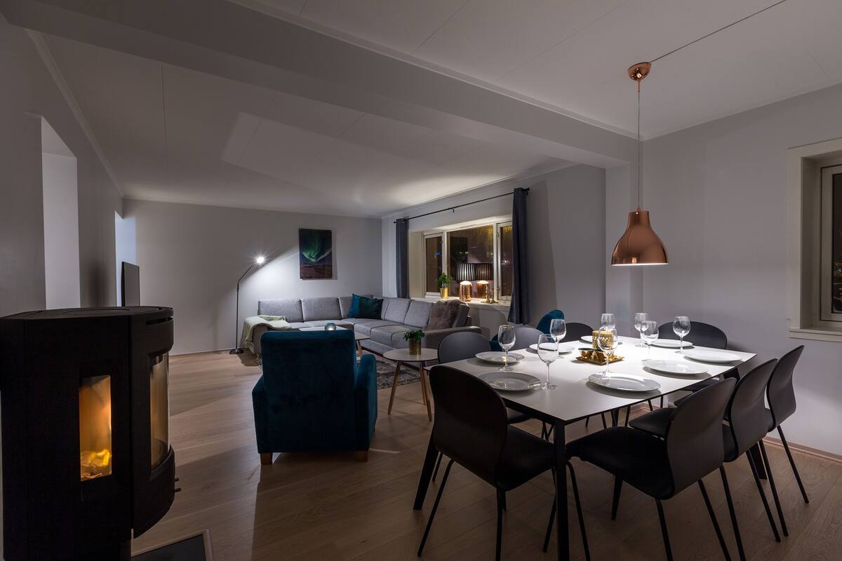 Uma sala de estar moderna com lareira, uma área de jantar com mesa posta para seis pessoas e um sofá com vista para o exterior.