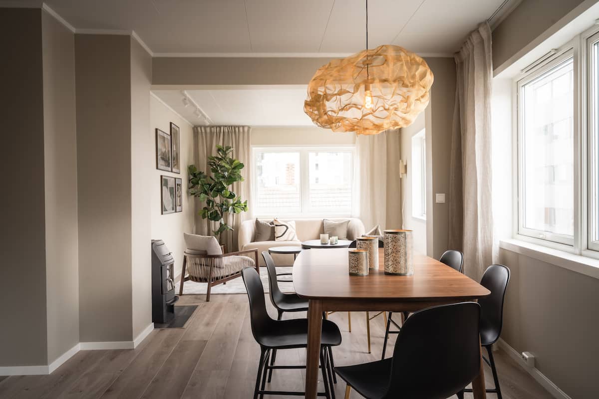 Uma área de refeições com mesa de madeira, cadeiras pretas e uma luminária pendente original. Há um sofá e uma planta ao fundo.