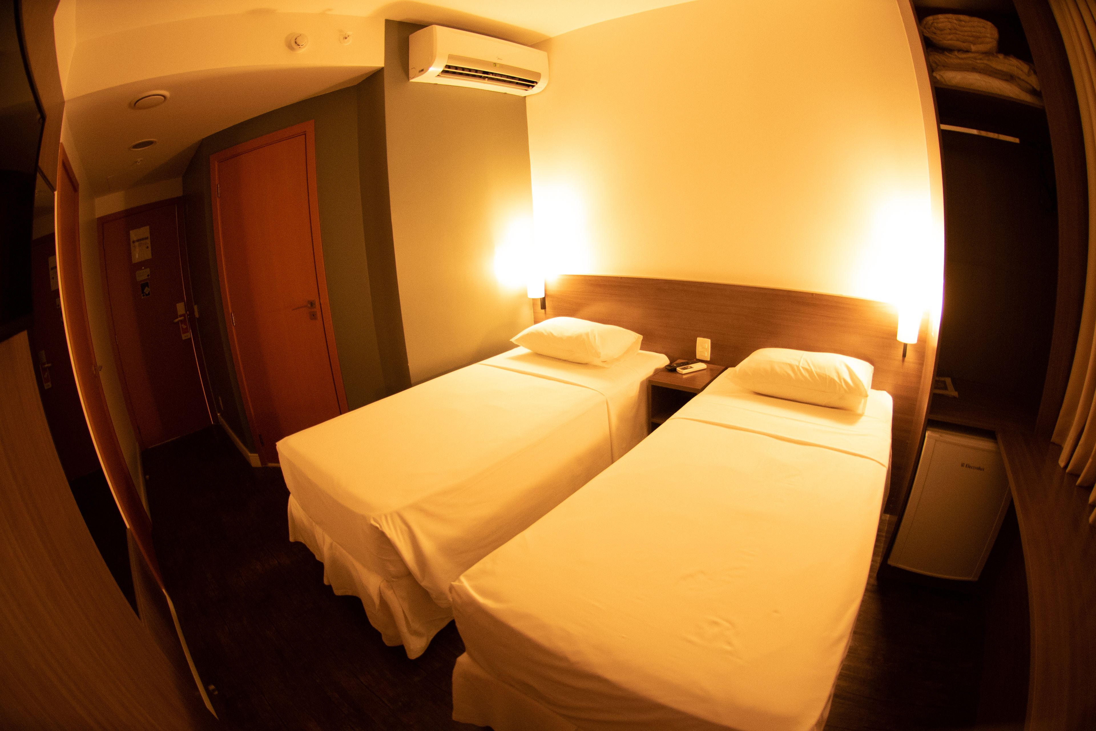 Quarto de hotel com duas camas de solteiro, cabeceira de madeira, mesa de cabeceira com abajur e unidade de ar condicionado.