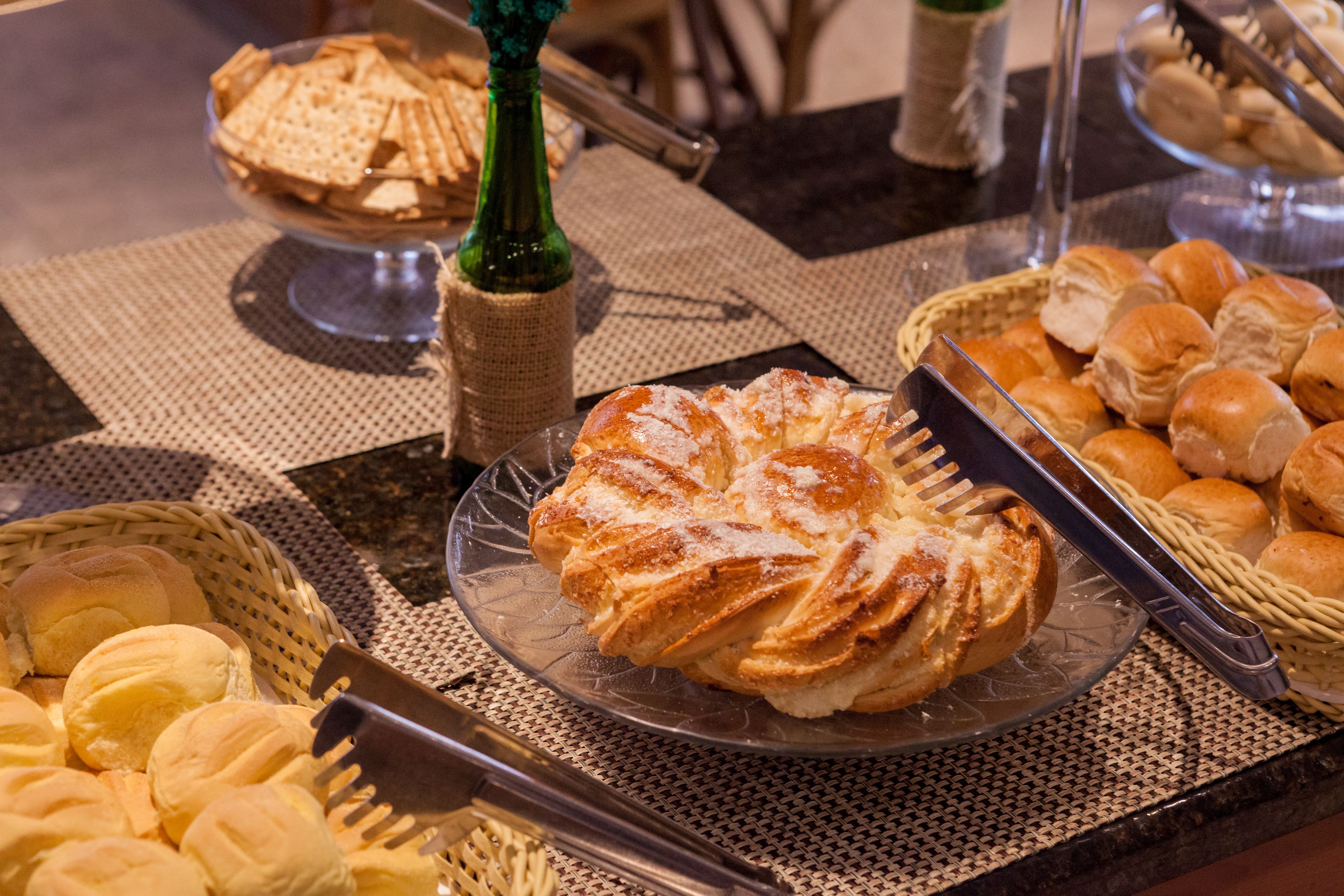 Uma mesa de buffet com diversos produtos de panificação, incluindo pães, doces e um bolo grande.