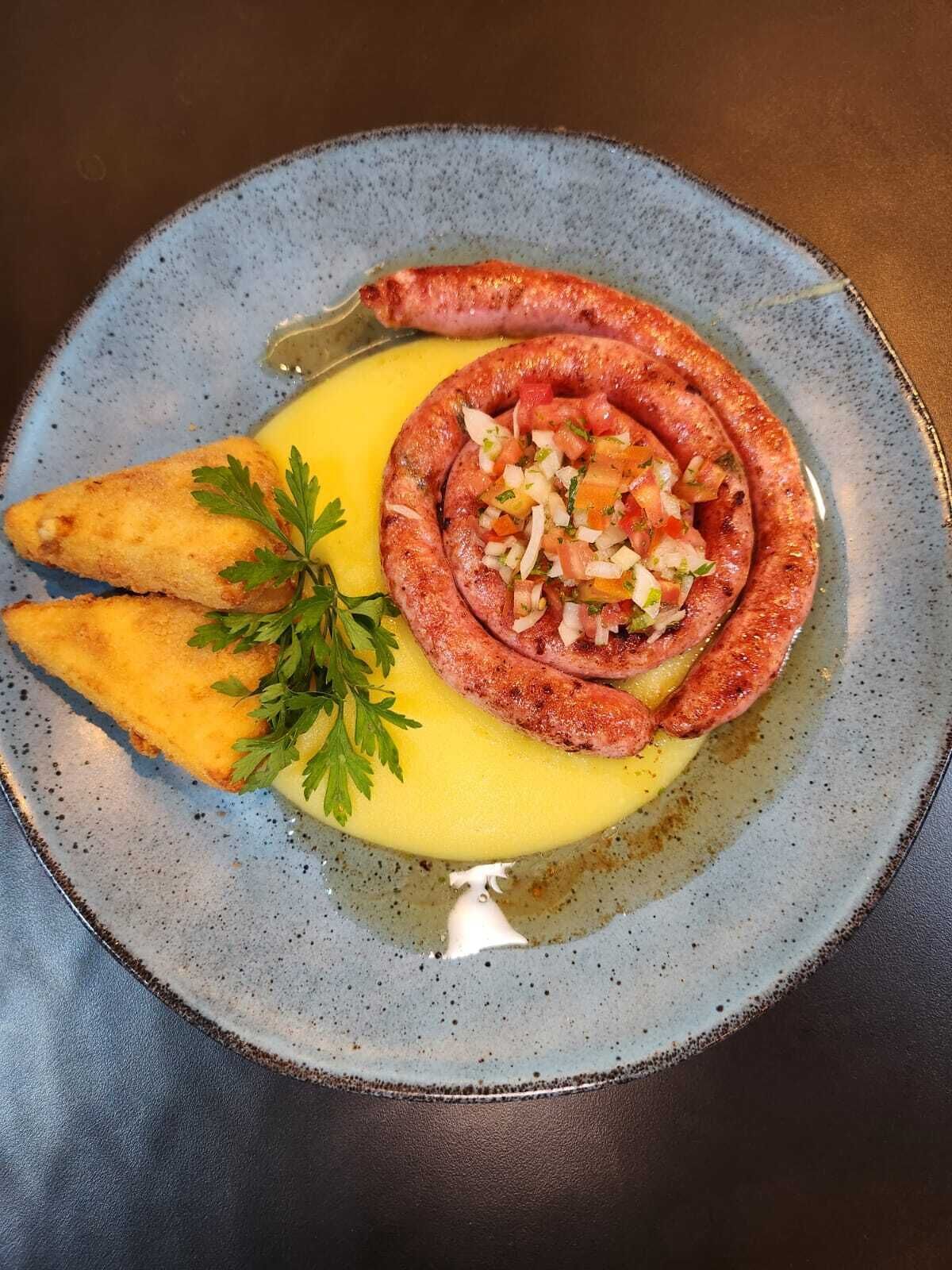 Um prato com uma espiral de salsicha cozida, um enfeite de salsa e dois itens fritos sobre um prato de cerâmica texturizada.