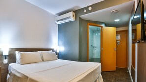 Quarto de hotel com cama, ar condicionado, mesa de cabeceira, luminária na parede e porta que dá para outro quarto.