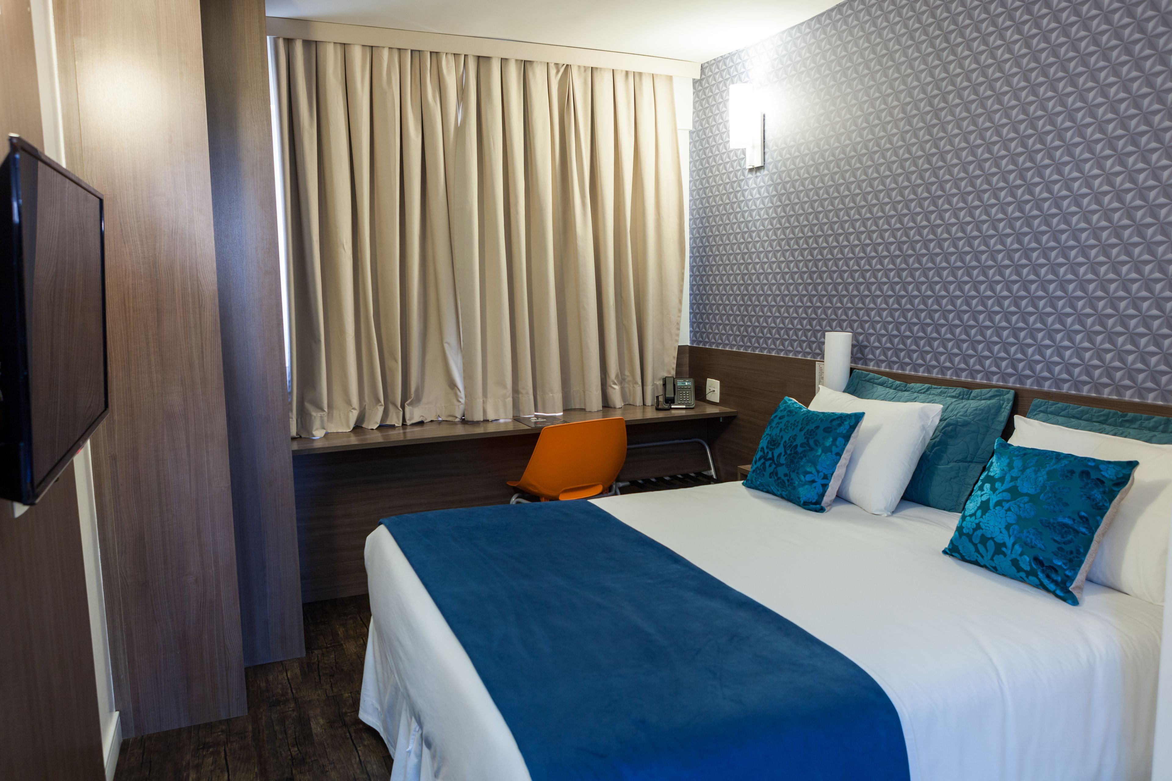 Quarto de hotel com cama, escrivaninha, cadeira laranja e televisão.