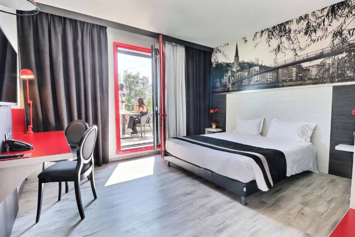 Quarto de hotel moderno com cama, escrivaninha, cadeira e vista para o exterior por uma porta de correr.
