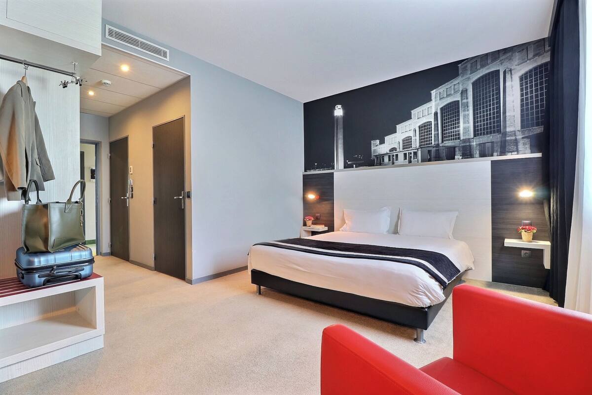 Quarto de hotel moderno com uma cama grande, uma cadeira vermelha, uma mala num armário e um mural na parede com a imagem de um edifício.