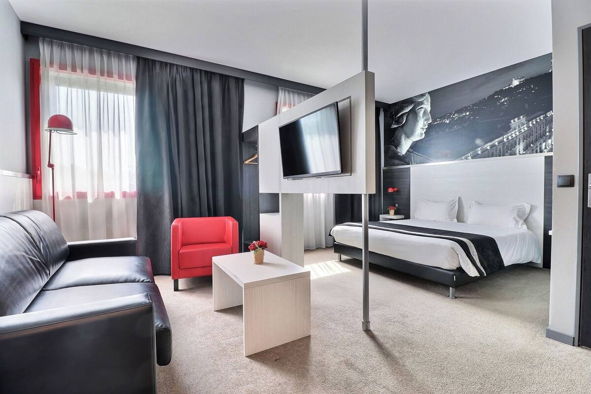 Quarto de hotel moderno com sofá de couro preto, uma cadeira vermelha, mesa de cabeceira branca, TV de tela plana fixada na parede, cama grande com roupa de cama branca e um mural com vista para a cidade.