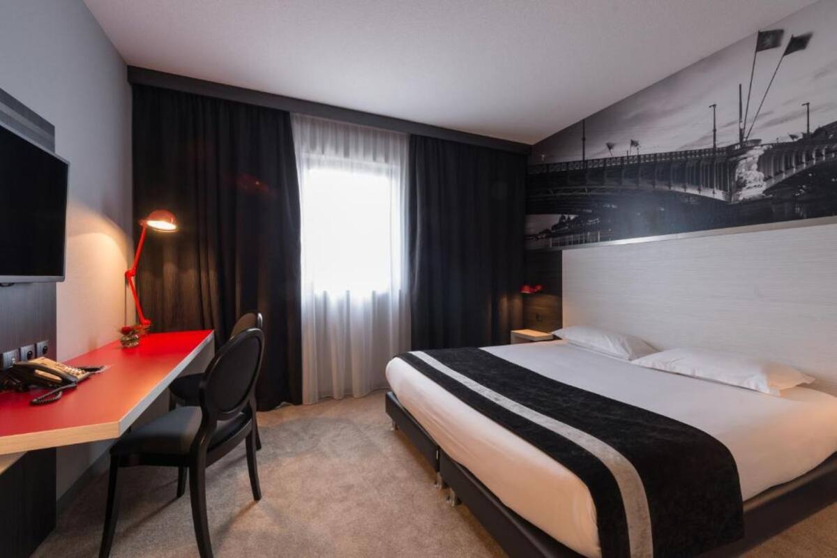 Quarto de hotel com uma cama, escrivaninha, cadeira, luminária e janela com cortinas.
