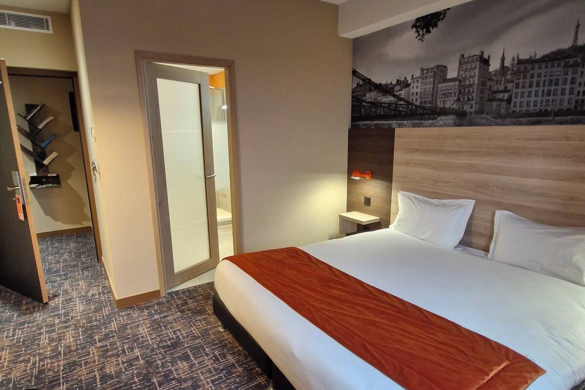 Quarto de hotel com cama grande, um mural da paisagem urbana, cabeceira de madeira e piso coberto por carpete.
