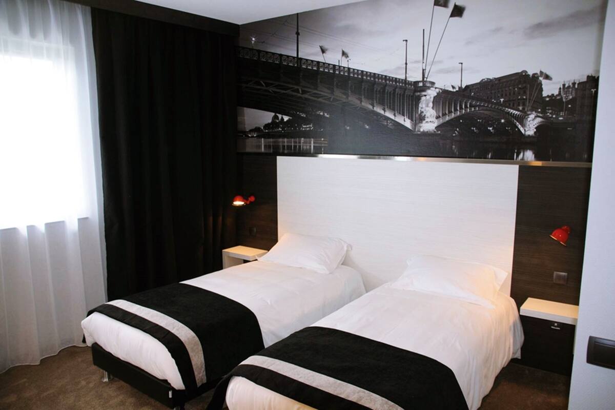 Quarto de hotel com duas camas, uma janela grande com cortinas finas, um mural com vista para a cidade na parede e uma mesa de cabeceira com um abajur vermelho.