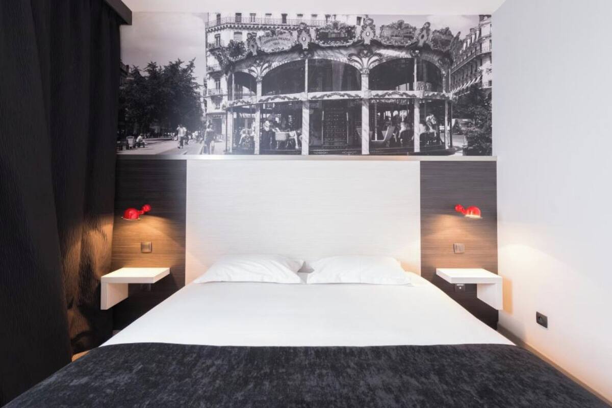 Quarto de hotel moderno com uma cama grande, duas mesas de cabeceira e um quadro fixado na parede retratando um edifício histórico.