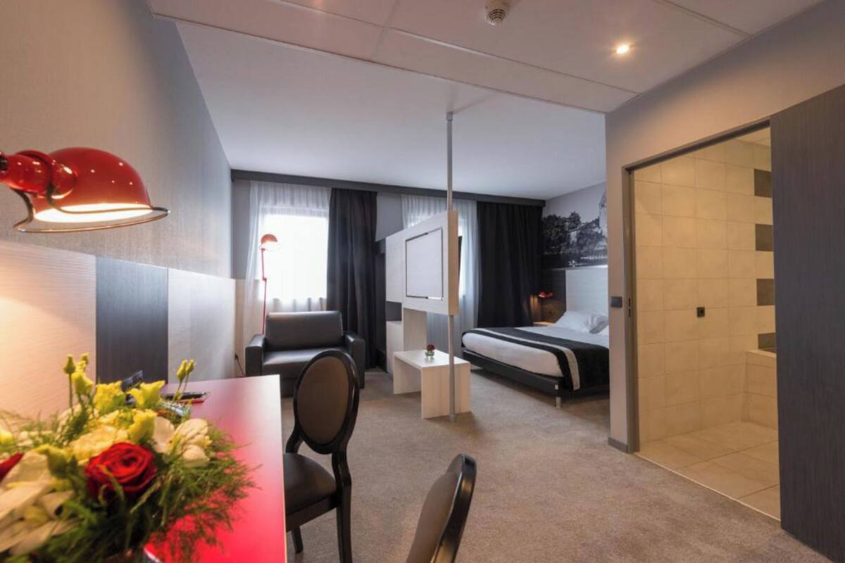 Quarto de hotel moderno com cama, escrivaninha, cadeira, televisão e área de chuveiro.