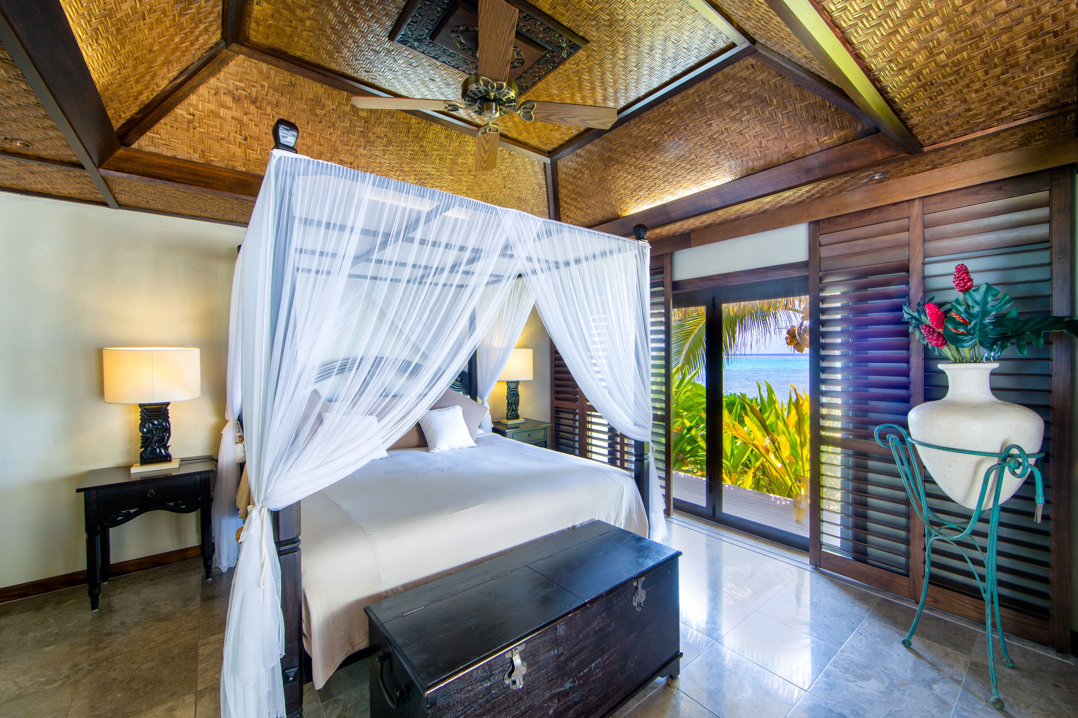 Quarto com cama sob dossel, mesa de cabeceira com abajur, baú de madeira e vista para o exterior.