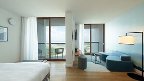 Quarto de hotel com uma cama, um sofá, uma mesinha, uma cadeira, uma luminária e vista para o exterior.
