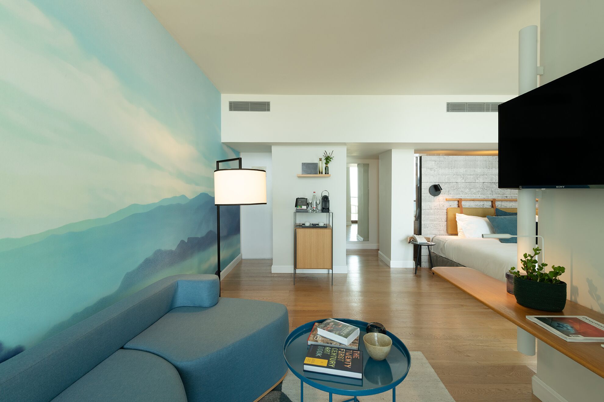 Quarto de hotel moderno com um sofá azul, uma mesa de centro redonda com livros, uma TV de tela plana e uma grande pintura de montanhas na parede.