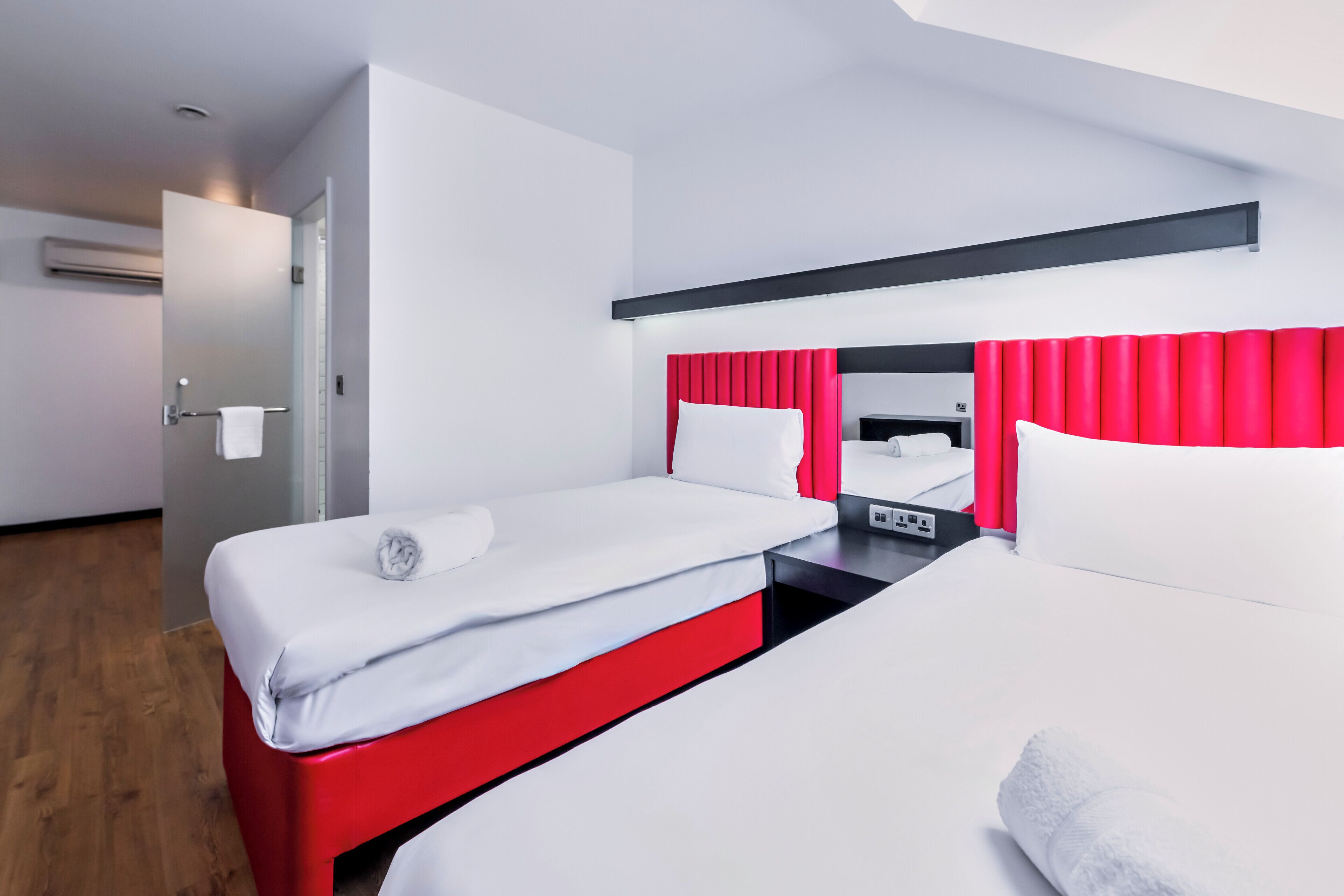 Quarto de hotel com duas camas, uma com base vermelha e roupa de cama branca, e um espelho grande sobre a cama.