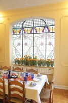 Uma sala de jantar com uma mesa posta para quatro pessoas, uma janela de vitrais com desenhos florais e vista para flores laranjas lá fora.