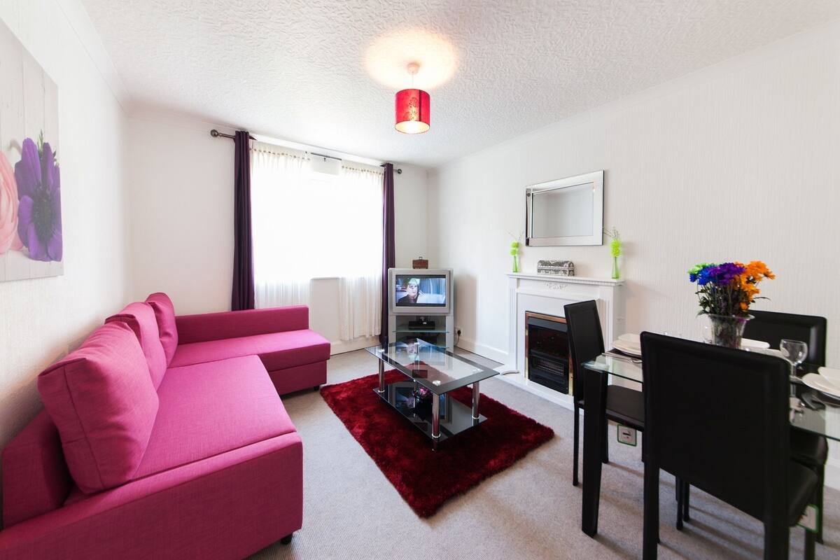 Uma sala de estar moderna com um sofá rosa, uma mesa de centro de vidro, uma lareira e um espaço de jantar com mesa e cadeiras.