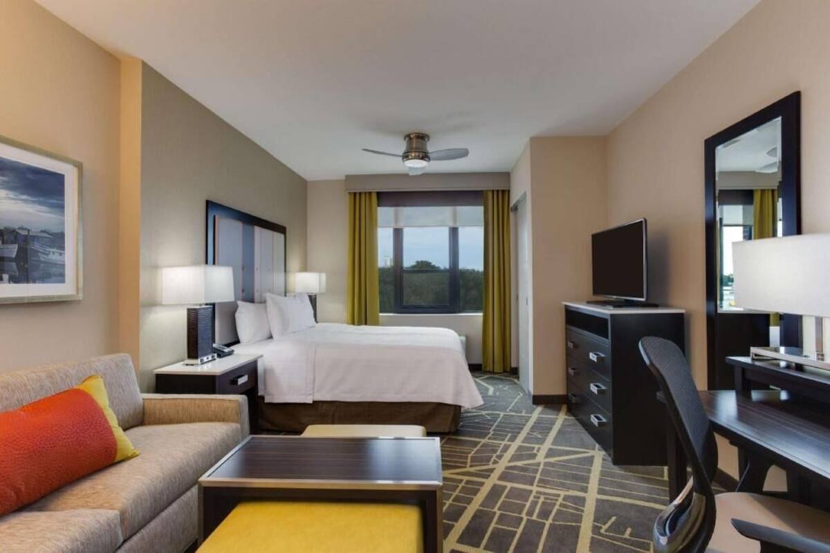 Quarto de hotel com cama, escrivaninha, cadeira, televisão, abajur, mesa de cabeceira, sofá e janela com cortinas.