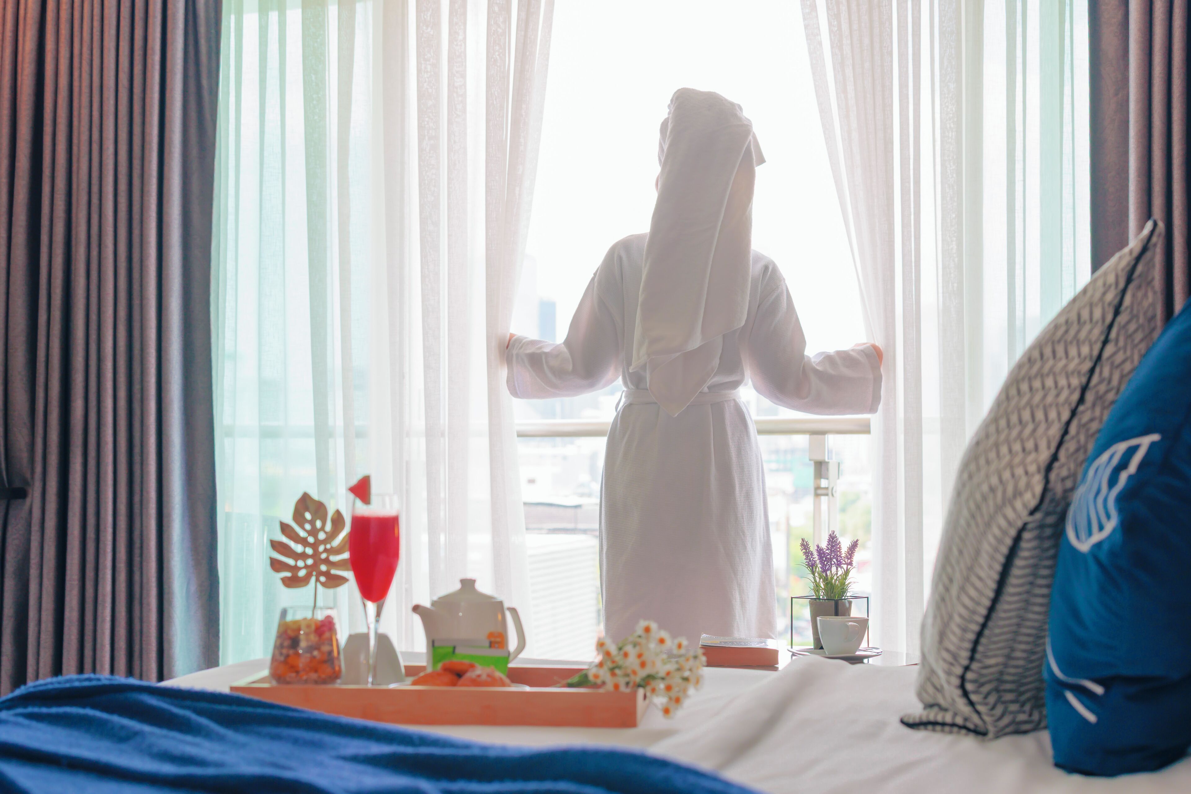 Quarto com uma cama, janela com cortinas, cafeteira, copo com bebida vermelha e bandeja com comida e flores.