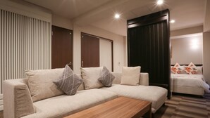 Quarto de hotel moderno com sofá, mesa de centro de madeira, cama com almofadas florais e cortina escura.