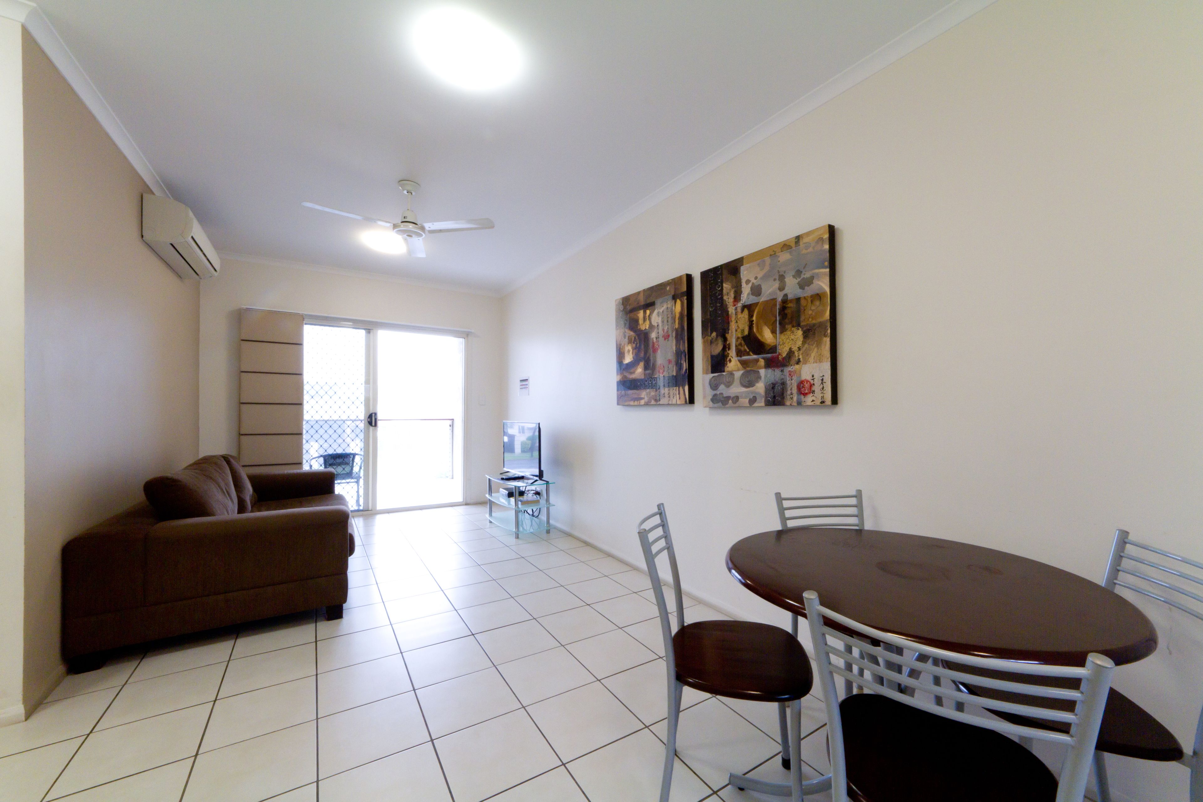 Uma sala de estar com um sofá marrom, uma mesa de jantar com quatro cadeiras e duas pinturas na parede.