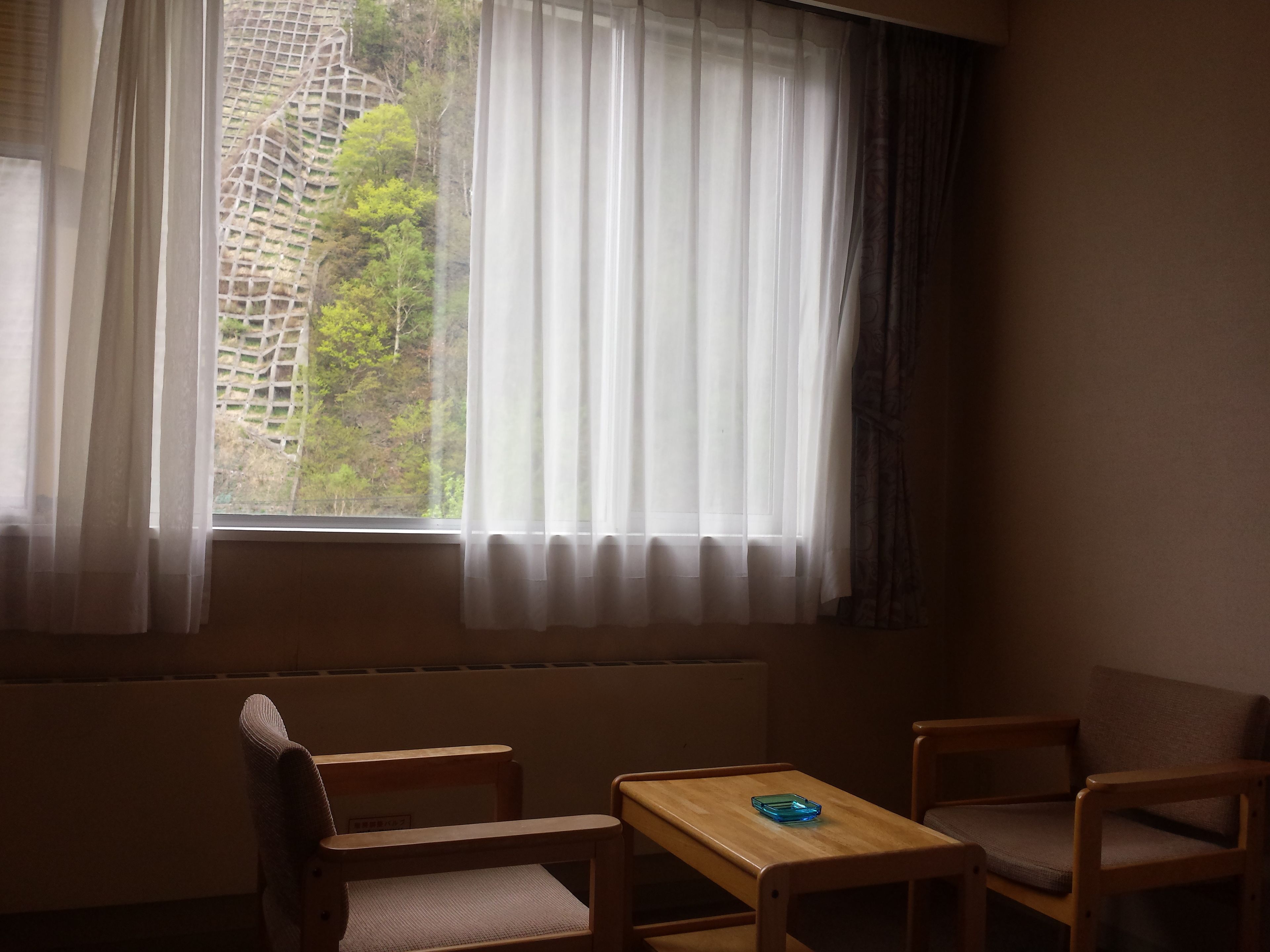 Um quarto com janela, duas cadeiras de madeira, uma mesa pequena de madeira e um objeto azul sobre a mesa.