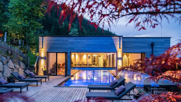 Uma casa moderna com piscina, cadeiras de descanso e vista para uma encosta arborizada.