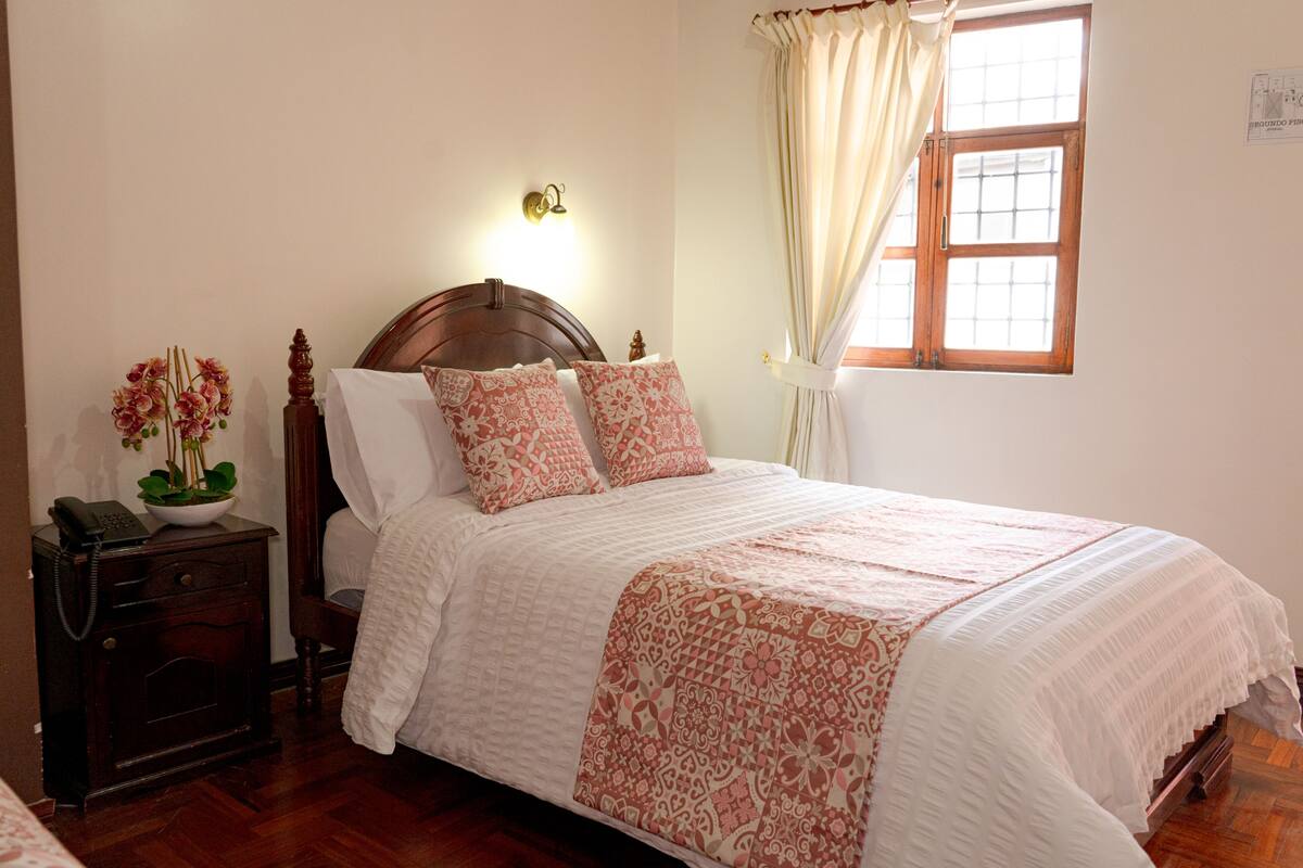 Cama bem arrumada com colcha branca e rosa estampada, mesa de cabeceira de madeira com um telefone e uma janela com cortinas.