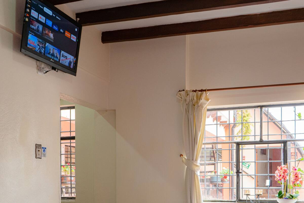 Quarto com duas camas, televisão na parede, janela com cortinas e vista para a rua.