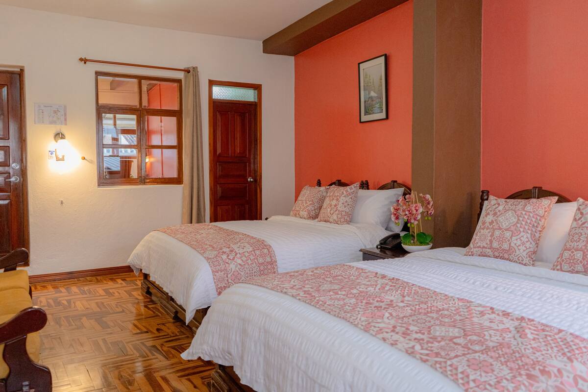 Quarto de hotel com duas camas, piso de madeira, porta de madeira, janela com cortinas e luminária fixada na parede.