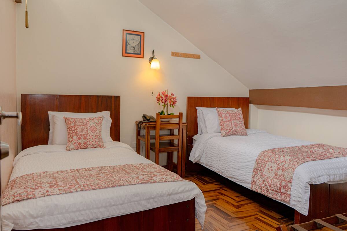 Quarto com duas camas, cabeceiras de madeira, uma cadeira de madeira, uma mesinha com um telefone, uma luminária na parede e um quadro emoldurado.