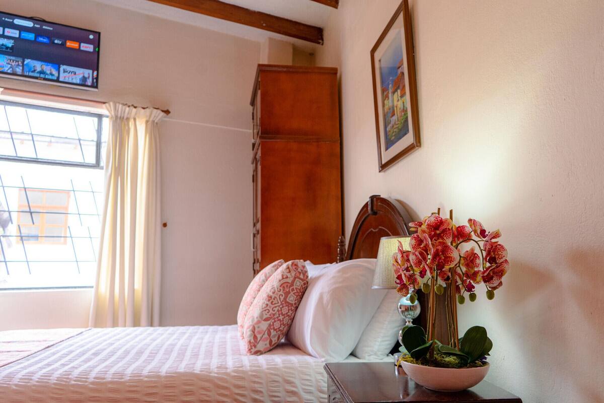 Quarto com cama, almofadas, televisão fixada na parede e janela com cortinas.