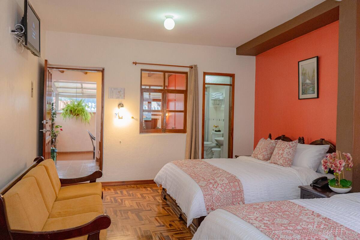 Quarto de hotel com duas camas, piso de madeira, sofá, banheiro visível por uma porta de vidro e uma TV fixada na parede.