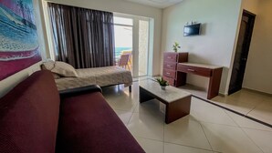 Quarto de hotel com cama, sofá, escrivaninha, televisão e vista para o oceano.