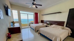 Quarto de hotel com duas camas, um ventilador de teto, uma escrivaninha com cadeira, uma mesinha com um vaso e vista para o mar através de uma porta de correr.