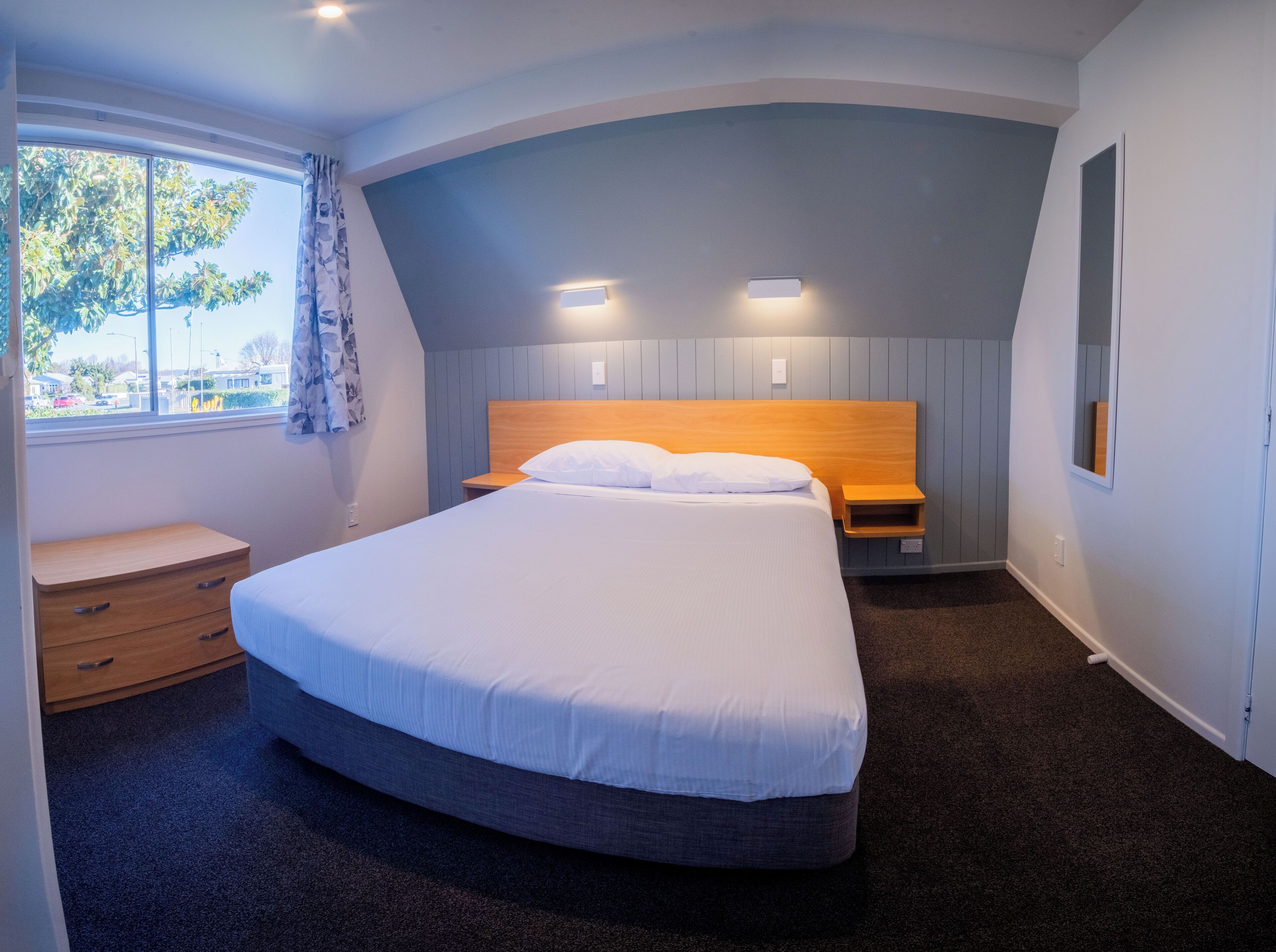 Um quarto com teto curvo, cama com roupa de cama branca, mesa de cabeceira de madeira, cômoda pequena de madeira e janela com cortinas.