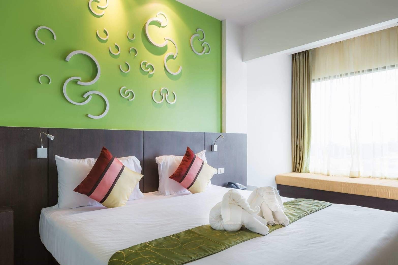 Quarto de hotel com parede verde decorativa, uma cama grande com roupa de cama branca, duas almofadas e um par de chinelos na cama.