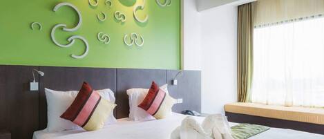 Quarto de hotel com parede verde decorativa, uma cama grande com roupa de cama branca, duas almofadas e um par de chinelos na cama.