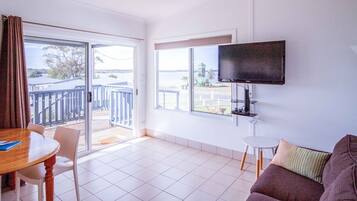 Quarto com televisão na parede, varanda com vista para o exterior, mesa de jantar com cadeiras e um sofá.